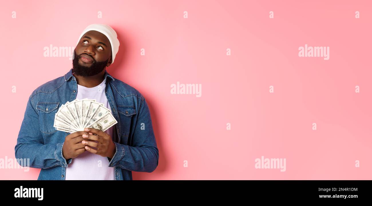 Dreamy african-american man thinking about shopping, holding money ...