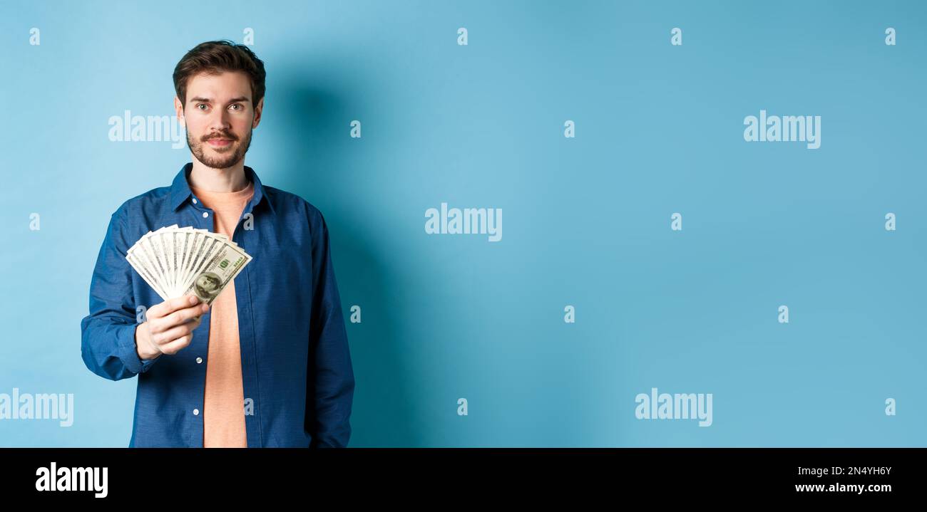 Happy modern guy holding money and smiling, standing on blue background ...