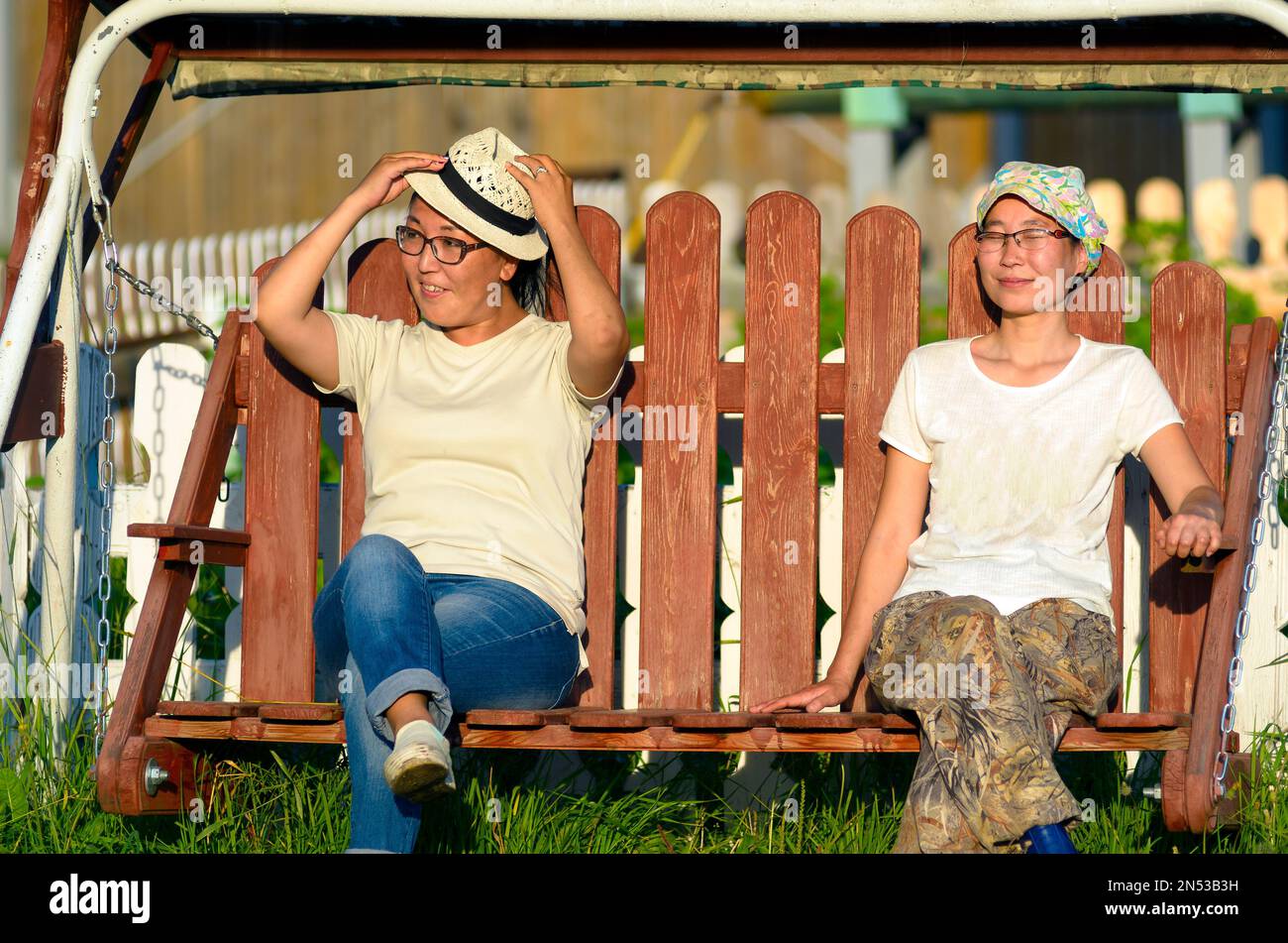 Two girls girlfriends Asian Yakuts relax on a swing next to flower beds ...