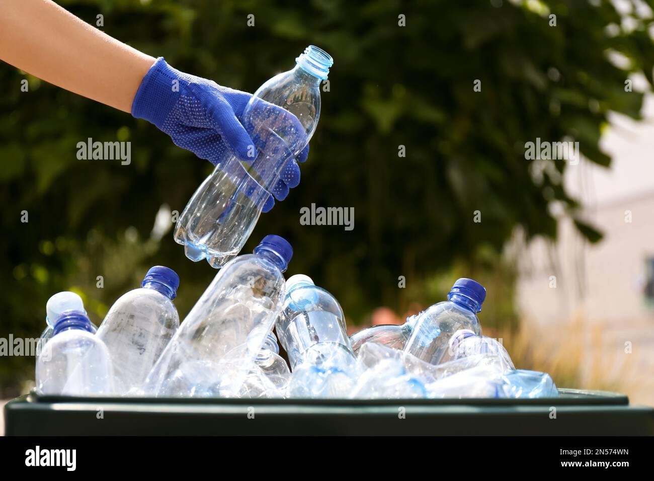 Woman in gloves putting used plastic bottle into trash bin outdoors ...