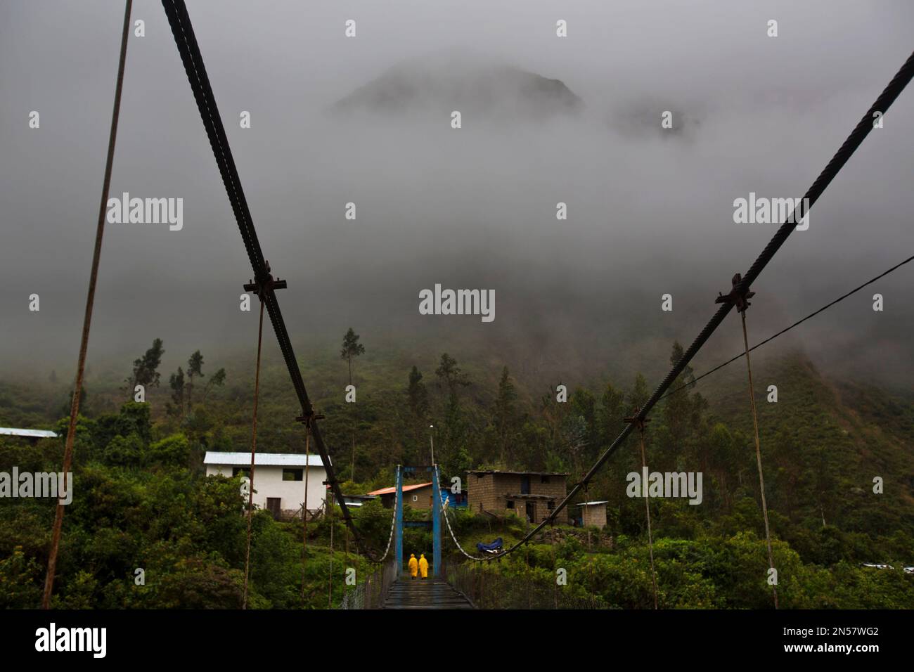 Two men in yellow rain coasts walk over a suspension bridge along the ...