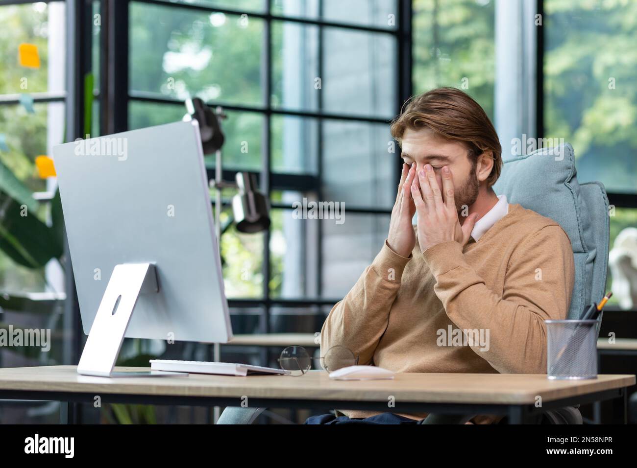 Tired office worker rubs eyes after long work at computer inside office, young blond businessman ...