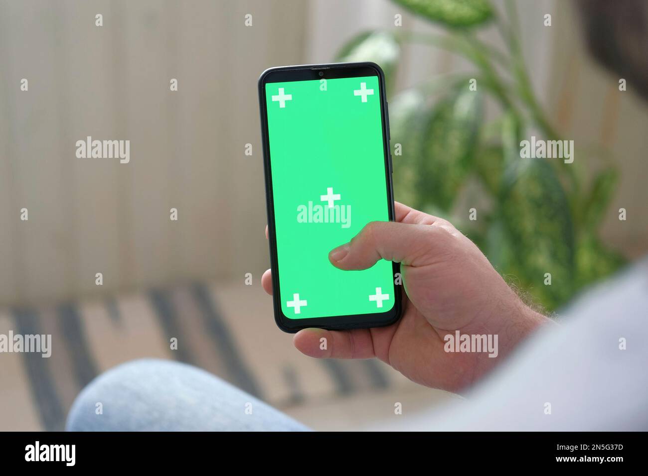 Young man sitting at home holding smartphone green mock-up screen in ...