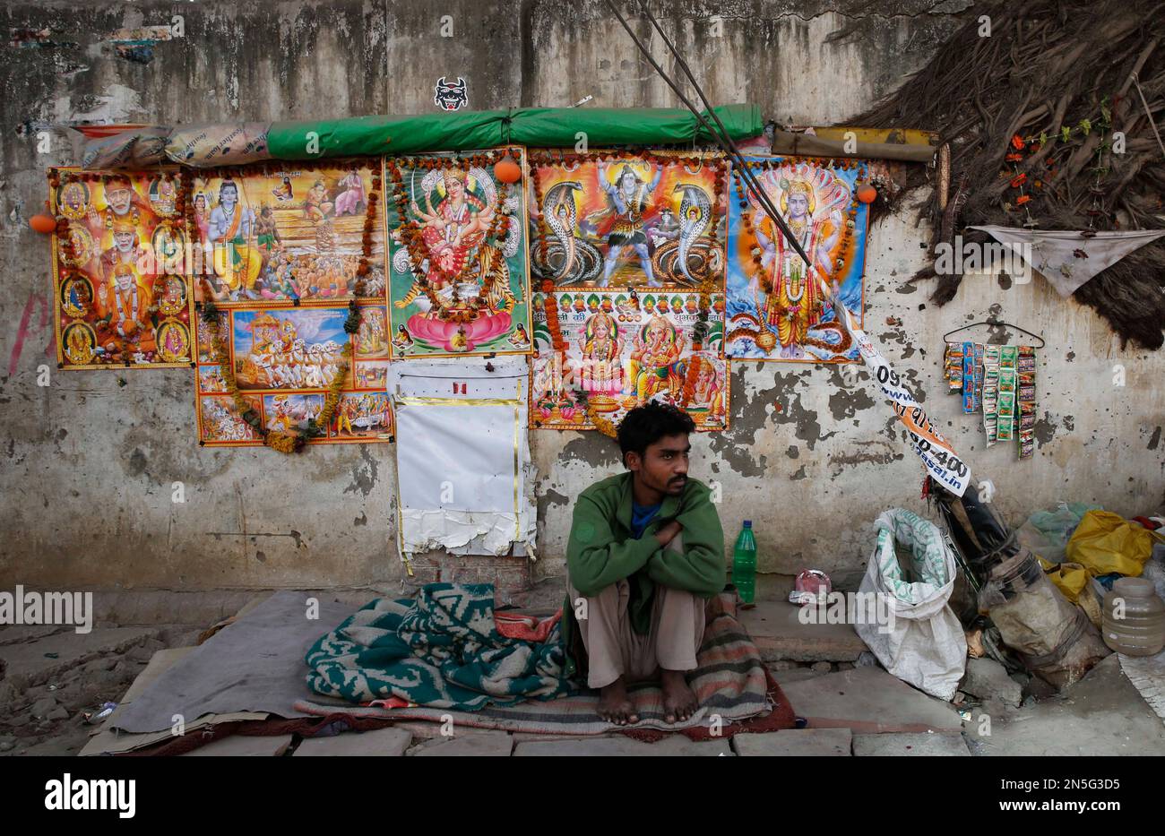 An Indian homeless man sits in front of portraits of Hindu gods and ...