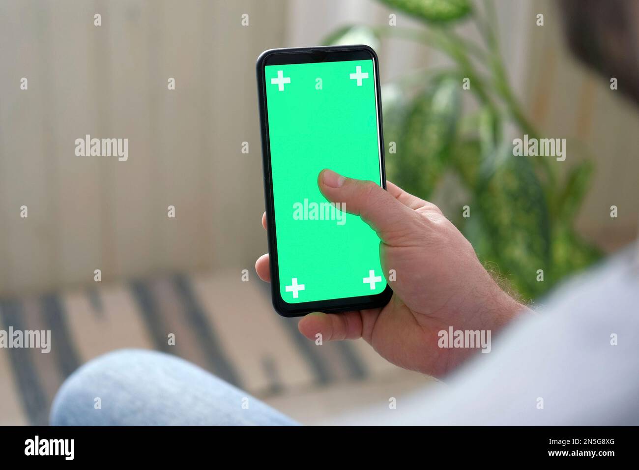 Young man sitting at home holding smartphone green mock-up screen in hand. Male person using ...