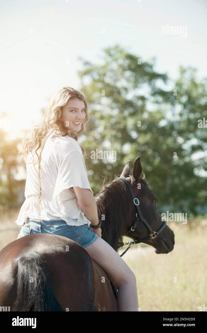 Young woman riding a horse in farm looking over shoulder and smiling ...