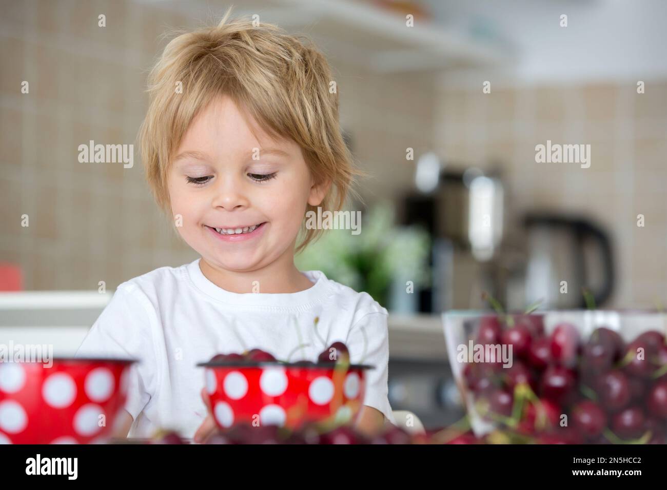 Cute blond child, toddler boy, eating cherries at home, having fun ...