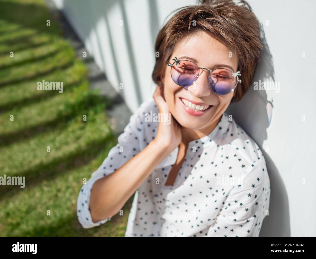 Smiling woman in colorful sunglasses has a rest on lawn in urban park ...