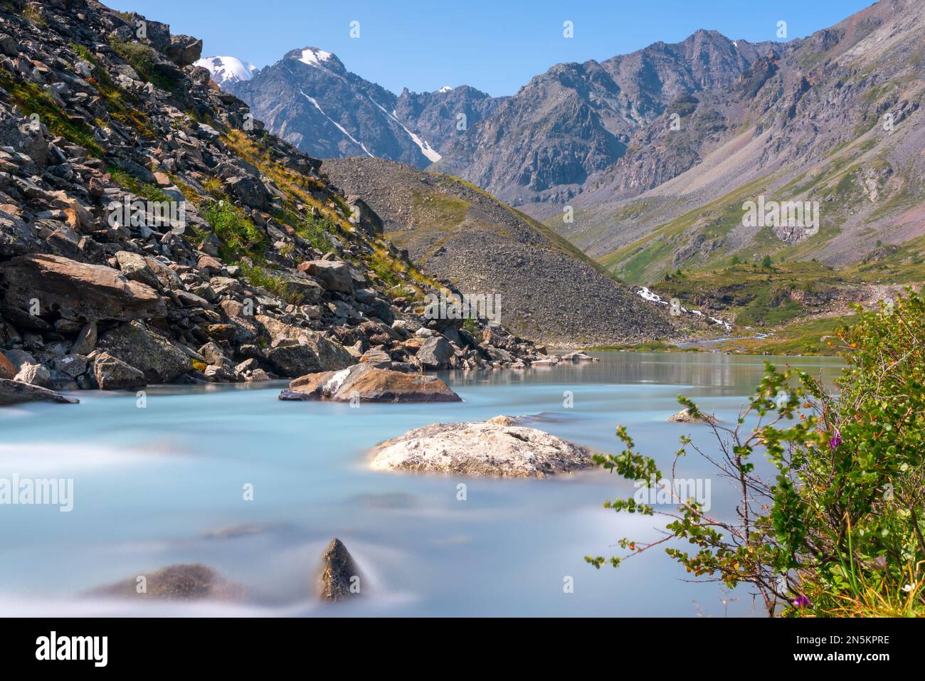An alpine stream flows over rocks near mountains down from a lake in ...