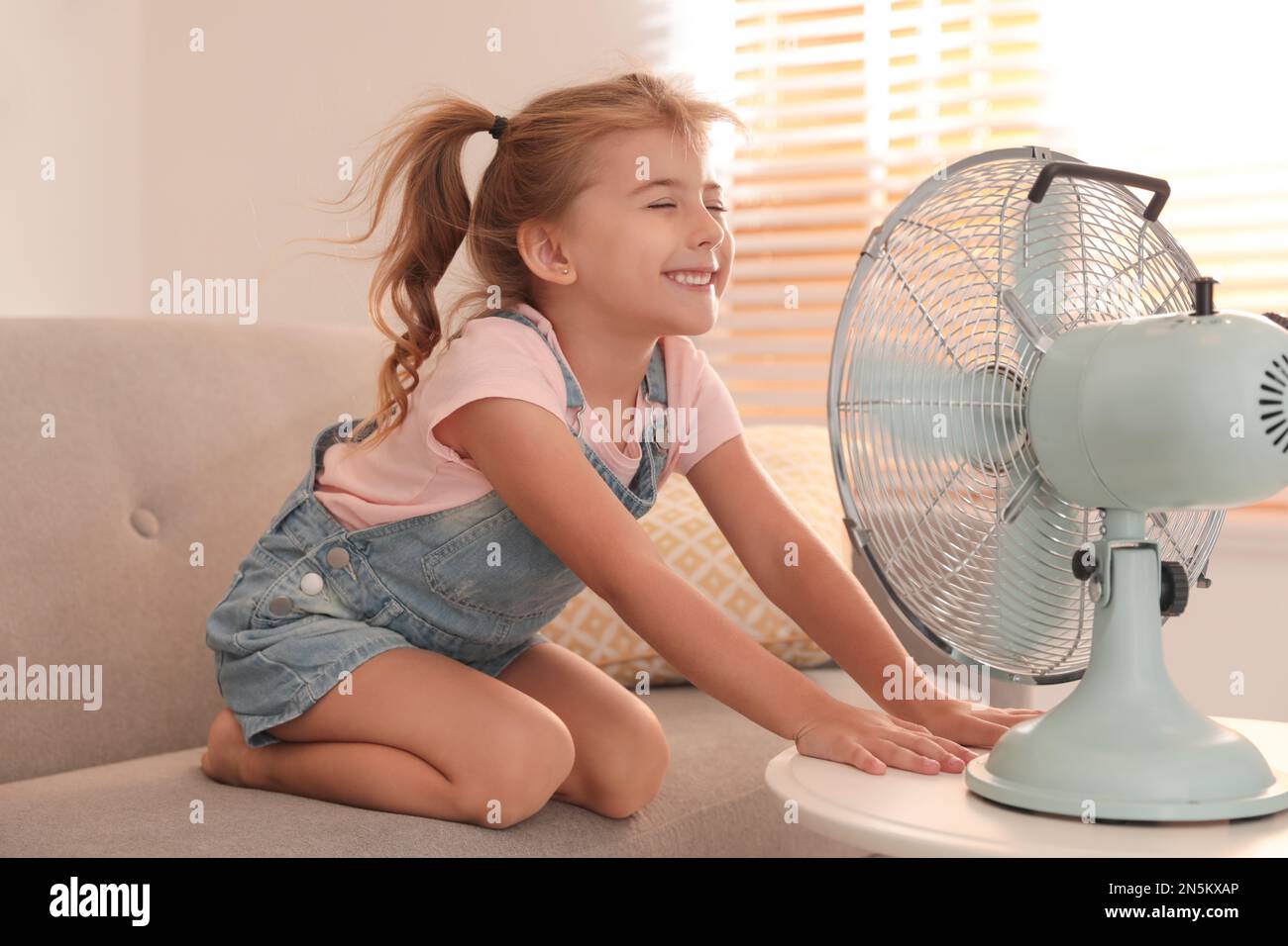 Little girl enjoying air flow from fan on sofa in living room. Summer ...