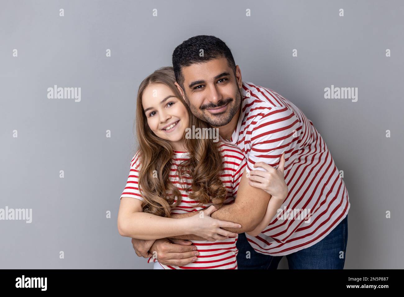 Portrait of loving happy father and daughter in striped T-shirts standing together, dad hugging ...