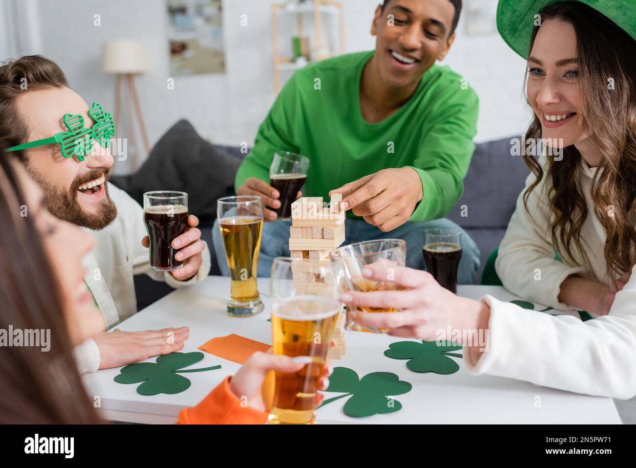 Cheerful women clinking alcohol drinks while interracial friends playing wood blocks game during ...