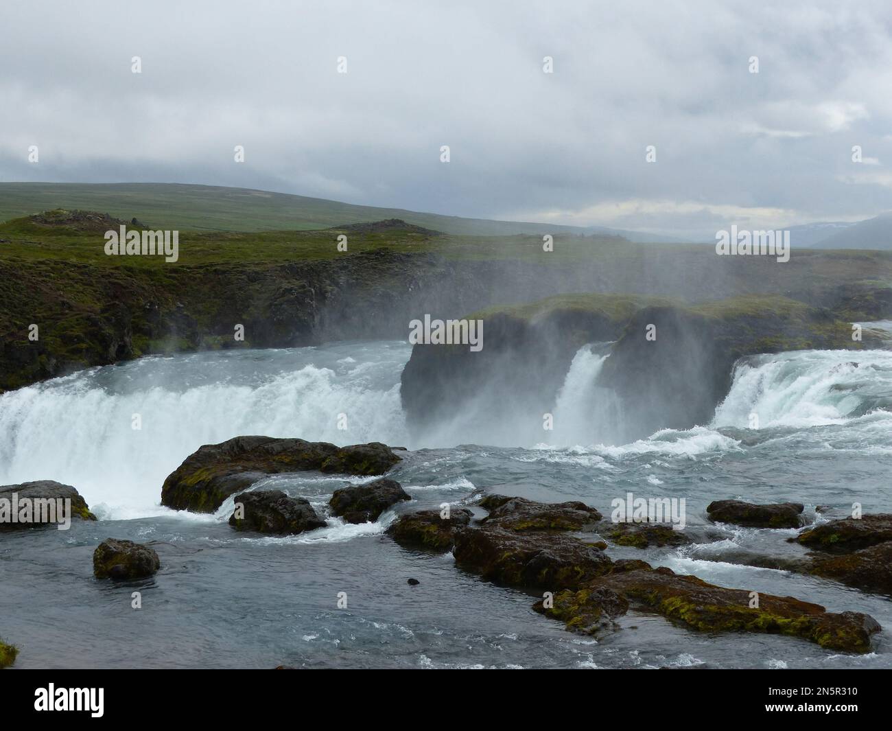 A landscape scene of Godafoss Waterfall with green hills and cloudy sky ...