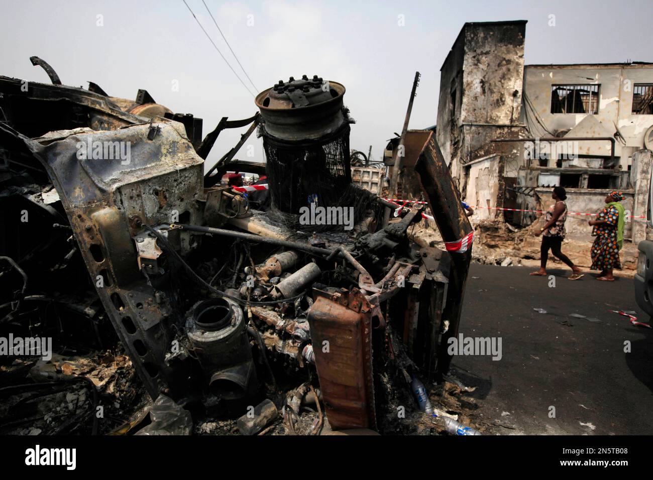 Women walk past a tanker that overturned and caused a large fuel fire ...