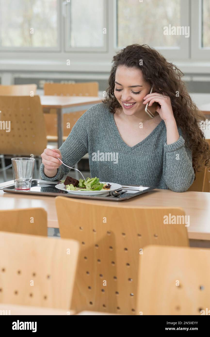 University student eating salad in canteen and talking on mobile phone ...