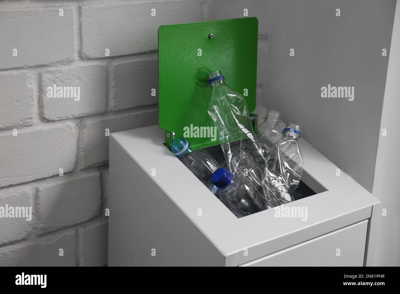 Many used plastic bottles in metal bin near white brick wall. Recycling ...
