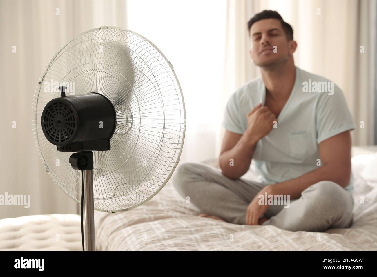 Man enjoying air flow from fan on bed in room, focus on device. Summer ...