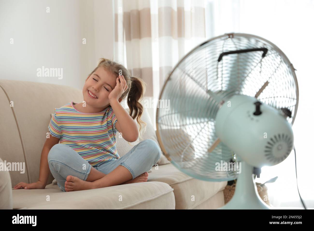 Little girl enjoying air flow from fan on sofa in living room. Summer ...