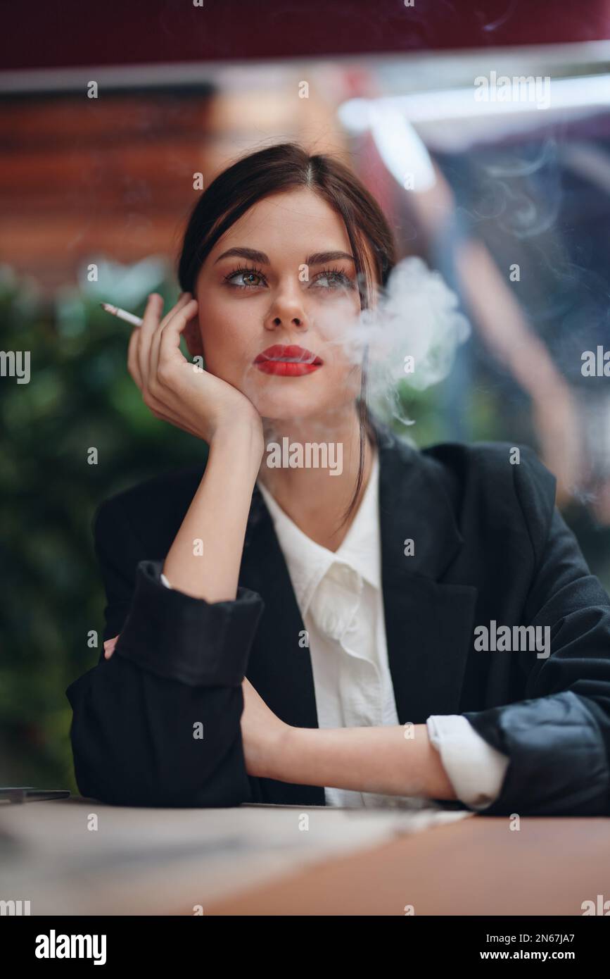 Stylish woman sits in a cafe at a table and smokes a cigarette ...