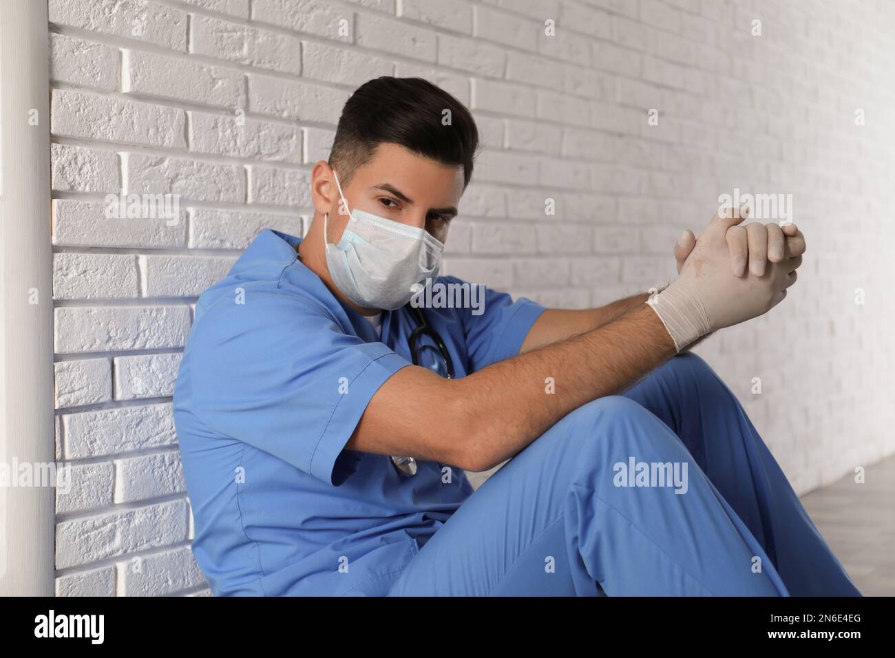 Sad doctor sitting near white brick wall indoors. Stress of health care ...
