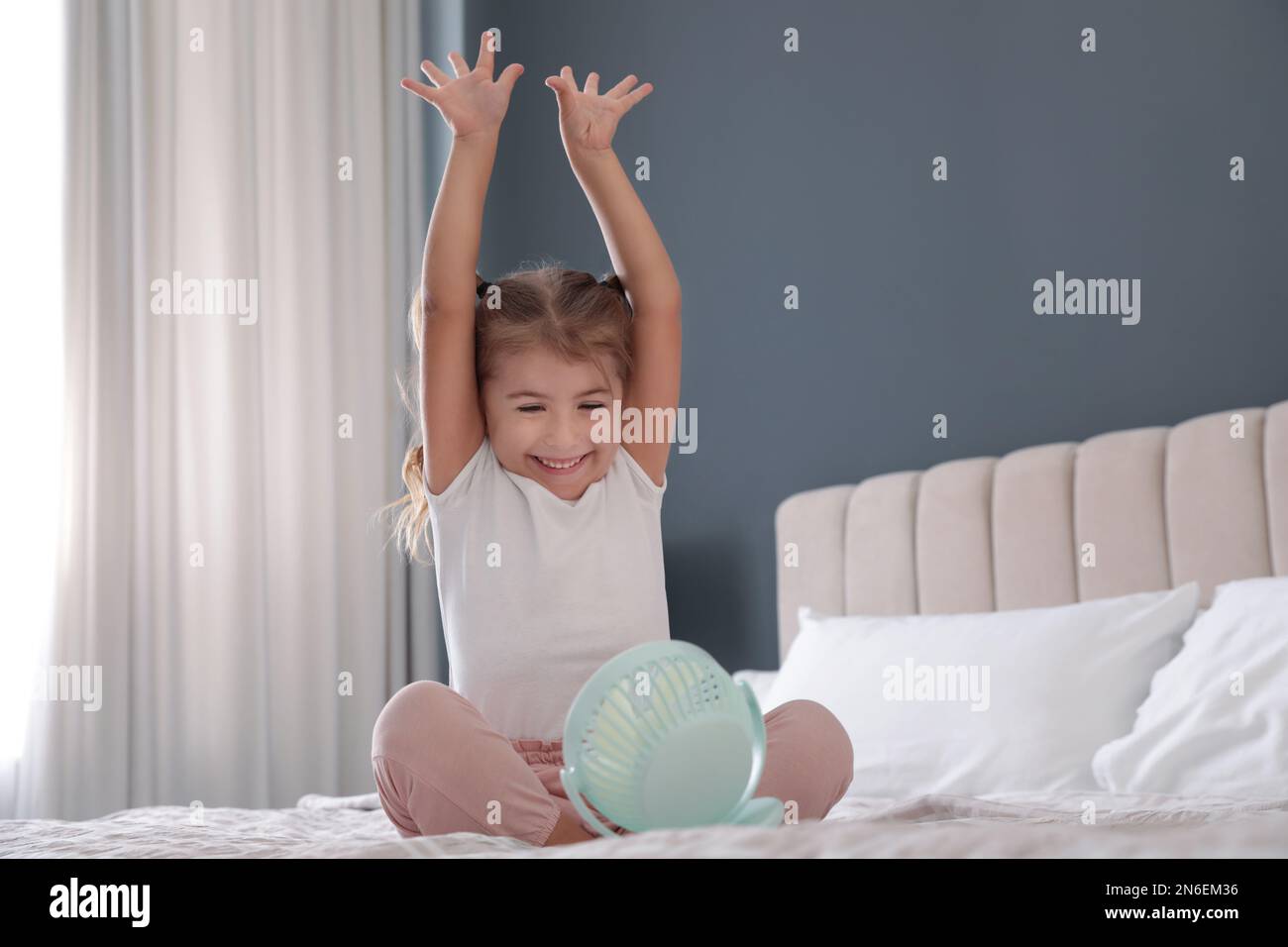 Little girl enjoying air flow from portable fan on bed in room. Summer ...