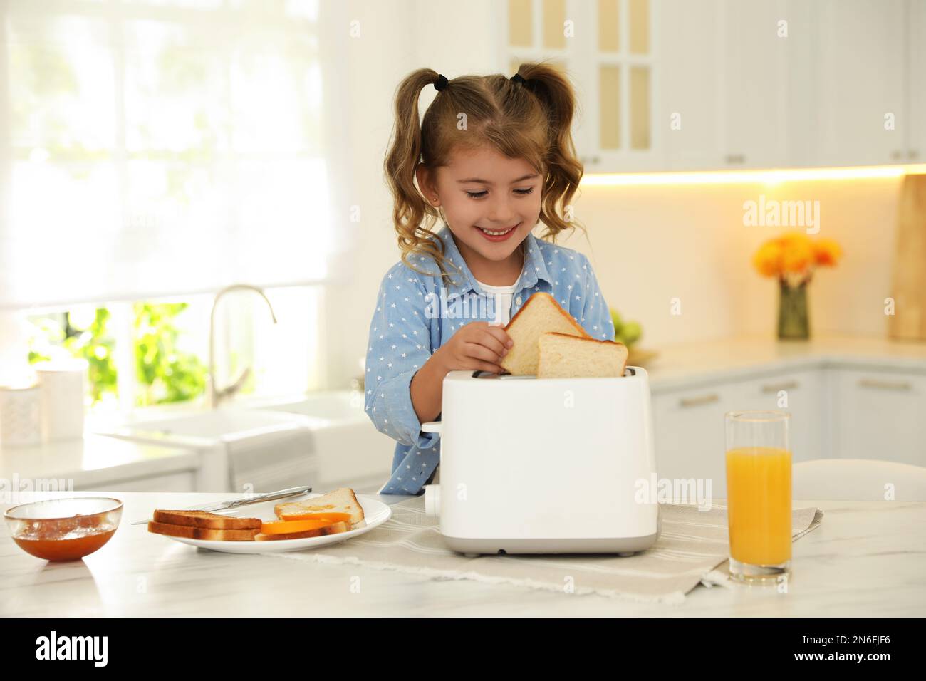 Cute little girl putting slice of bread into toaster at table in ...