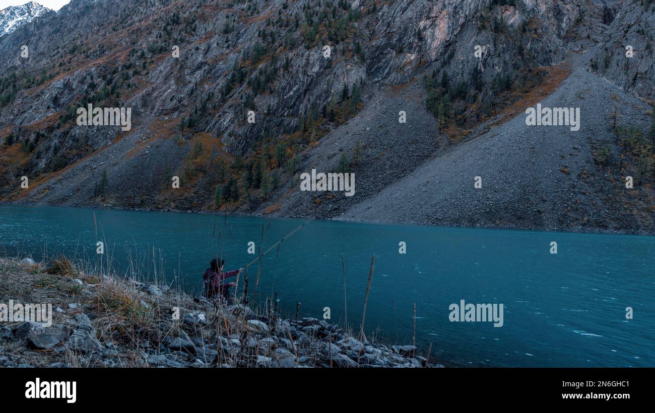 A girl fisherman sits on the shore of the alpine lake Shavlinskoye and ...
