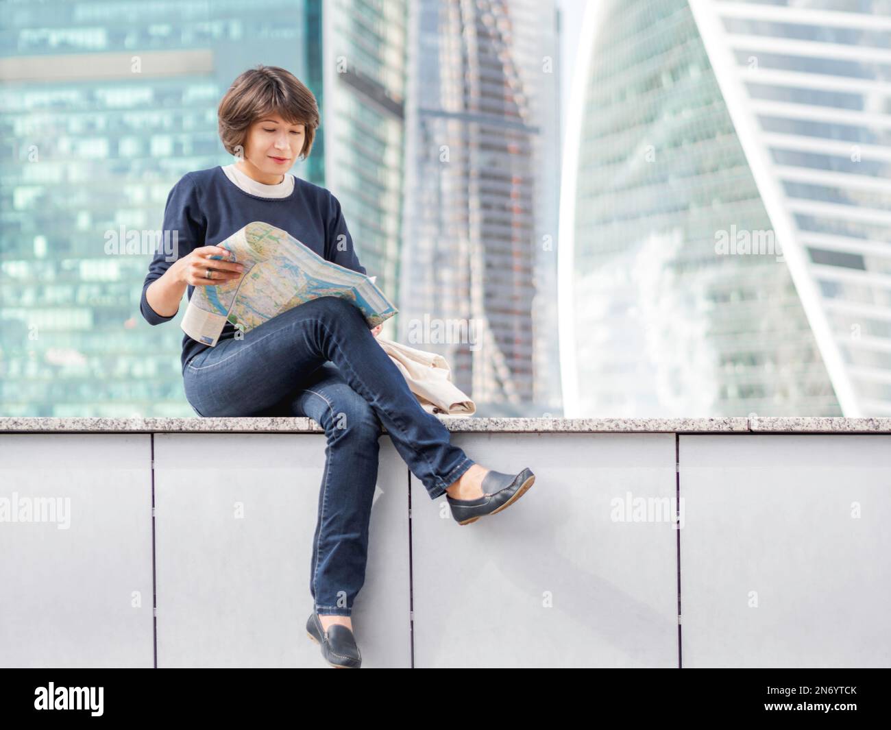 Smiling woman is reading paper map sitting on background of buildings ...