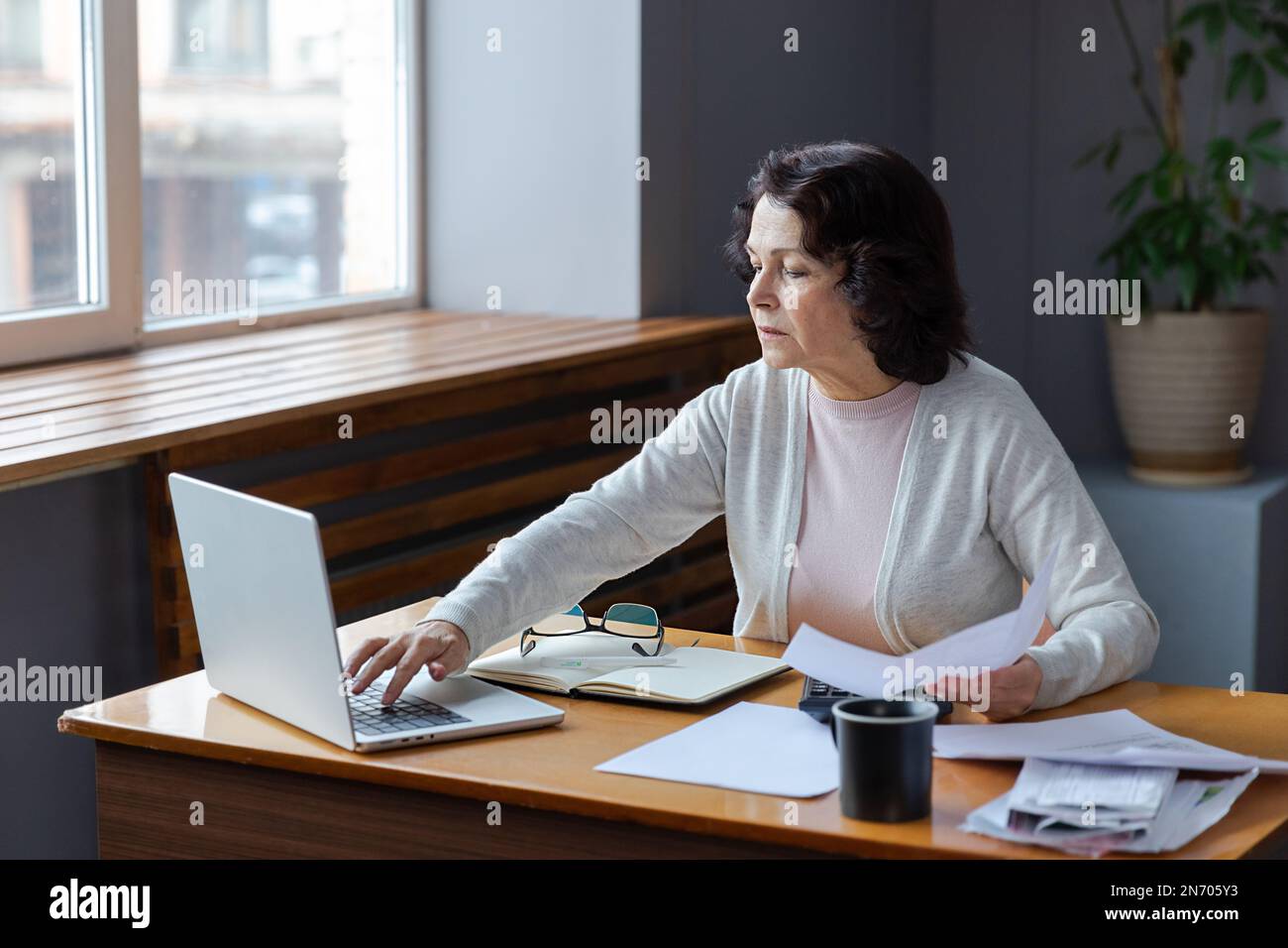 Middle aged senior woman sit with laptop and paper document. Pensive ...