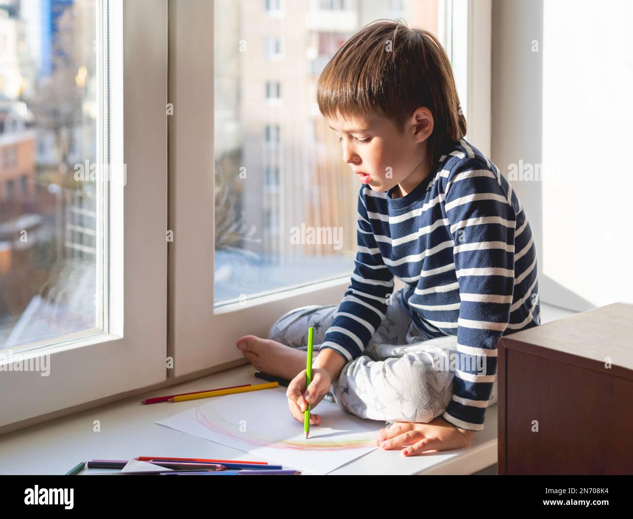 Little boy sits on window sill and draws rainbow with colored pencils ...