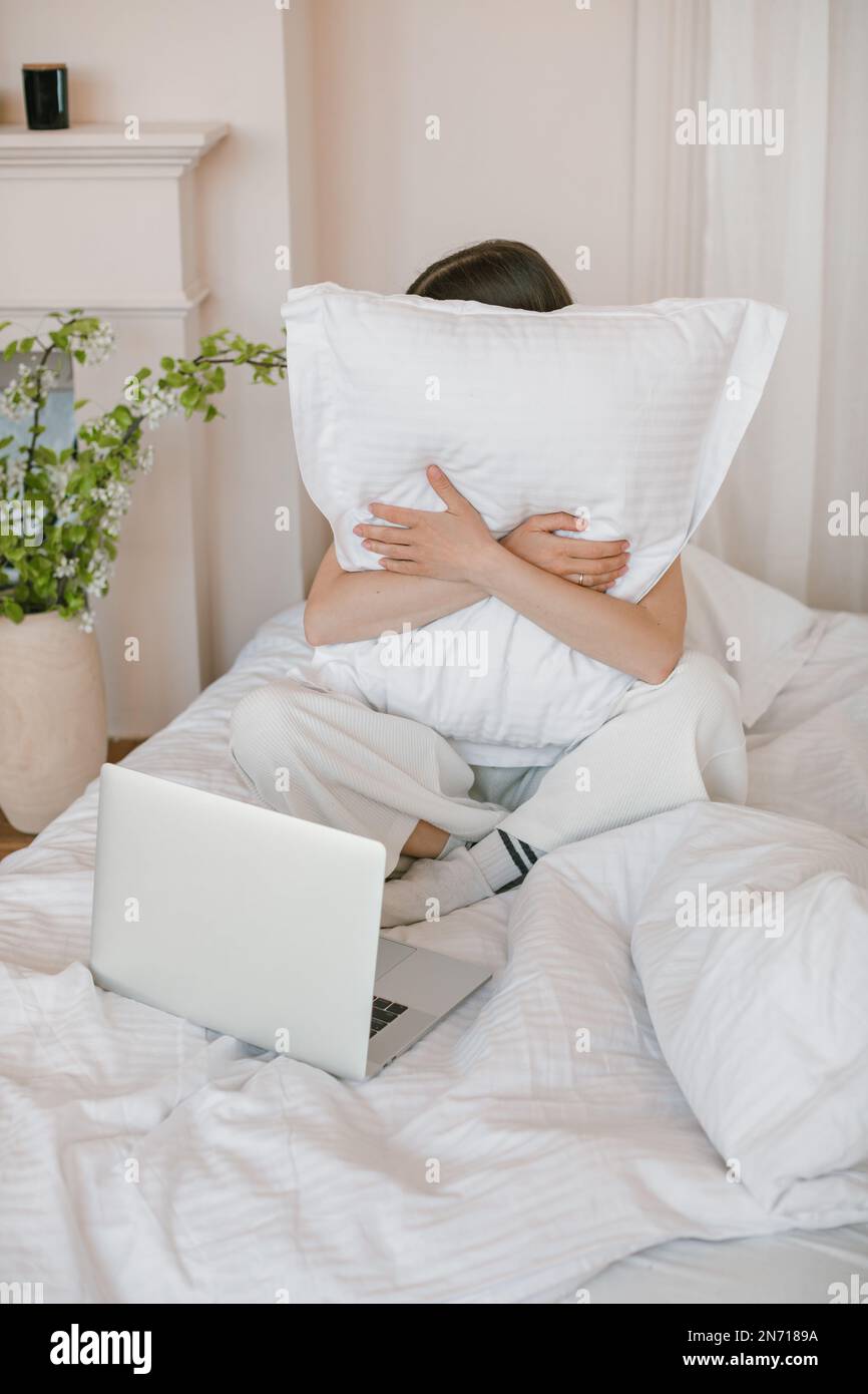 Woman sitting in bed in front of her laptop computer cuddling a pillow ...