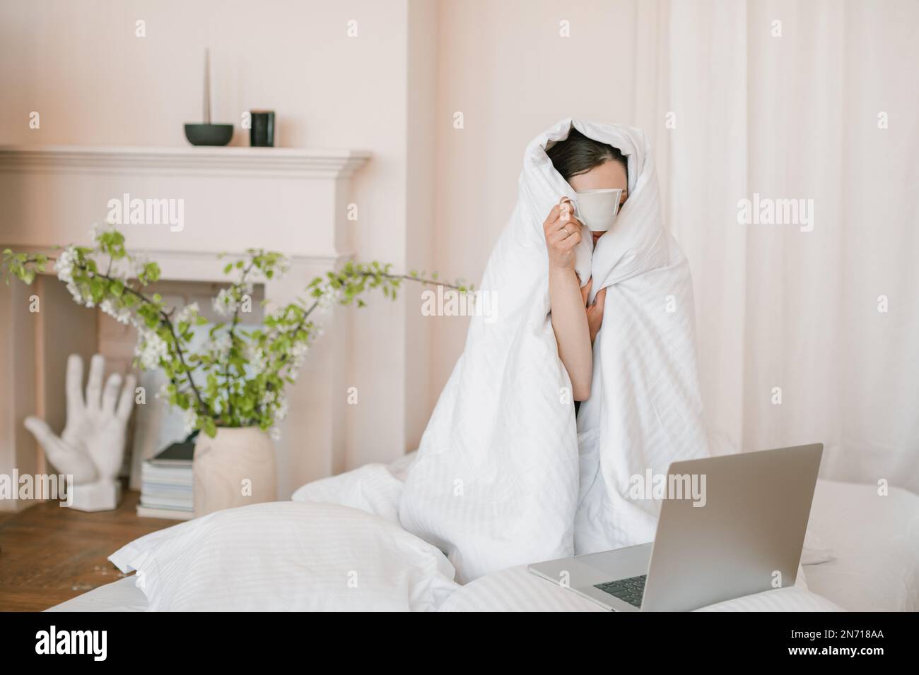 Woman wrapped in a duvet sitting in bed in front of a laptop computer ...