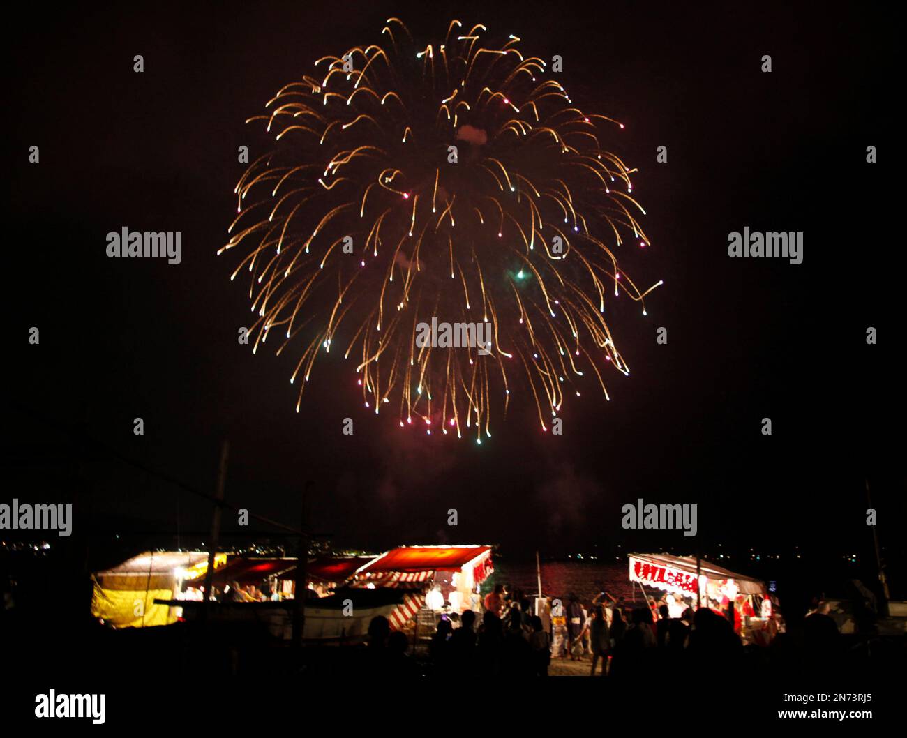 A firework explodes over sidewalk stalls on a beach during a summer ...