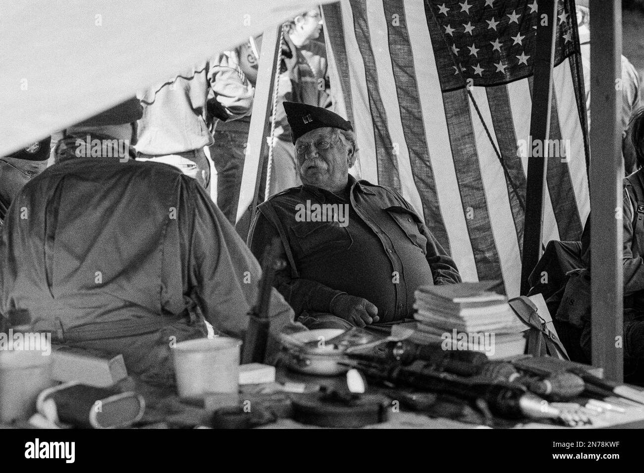 A weathered WWII Captain seated at a desk under a with an American flag ...