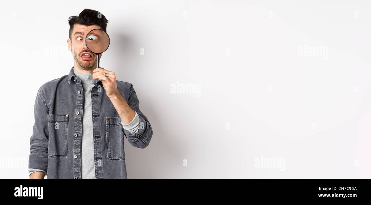 Funny young man squinting, showing faces with magnifying glass ...