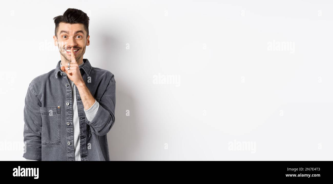 Happy guy shushing at camera, smiling with finger on lips, asking to ...