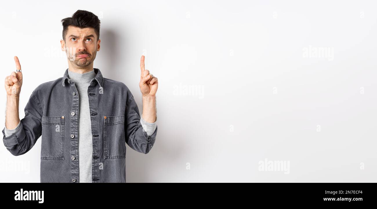 Sad frowning guy pointing and looking up, standing hesitant on white ...