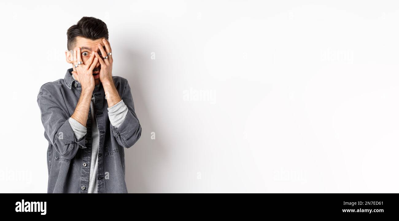 Scared young man peeking through fingers with nervous face, staring at ...