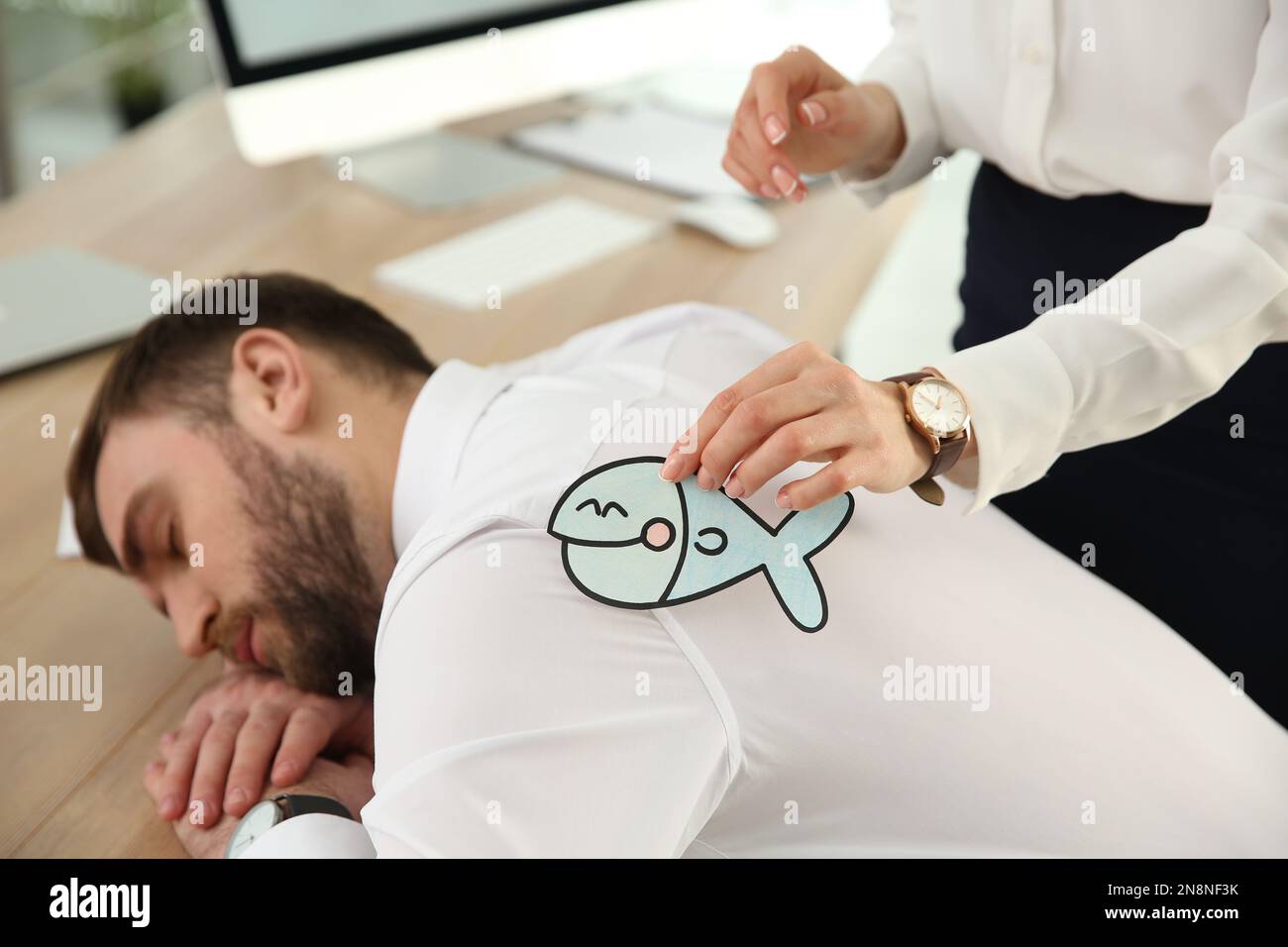 Young woman sticking paper fish to colleague's back while he sleeping ...