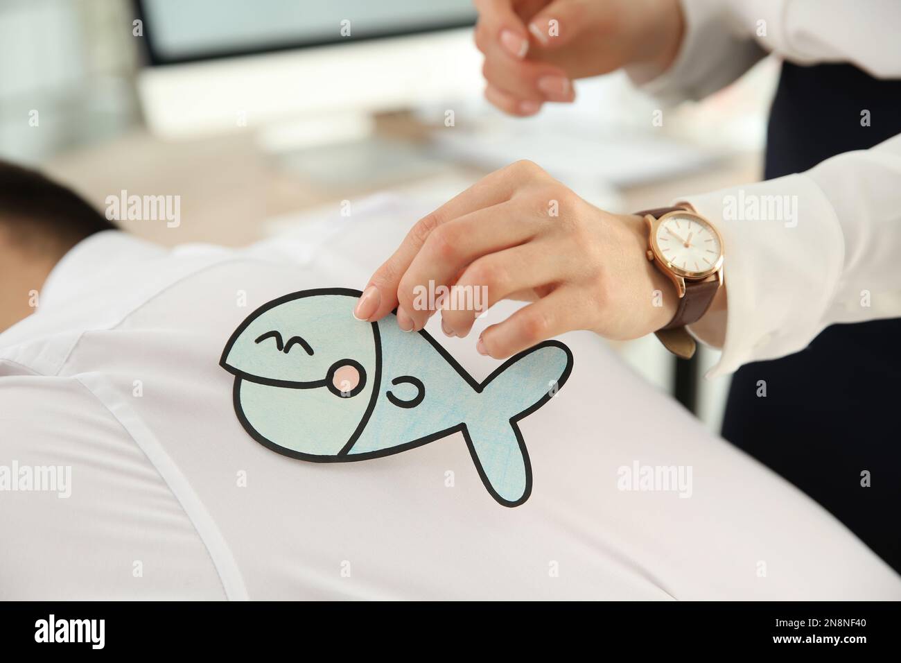 Young woman sticking paper fish to colleague's back in office, closeup ...