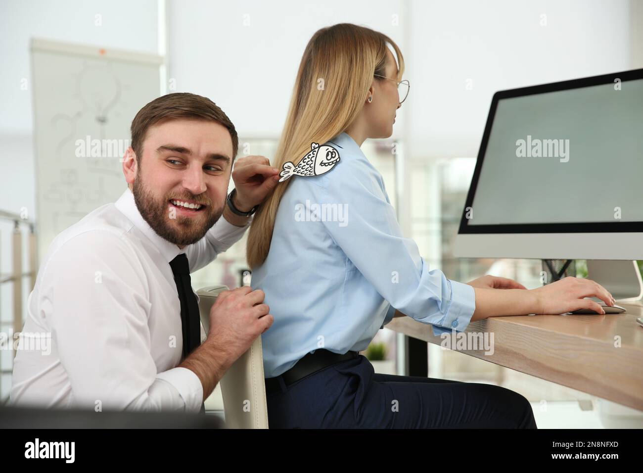 Young man sticking paper fish to colleague's back in office. Funny joke ...
