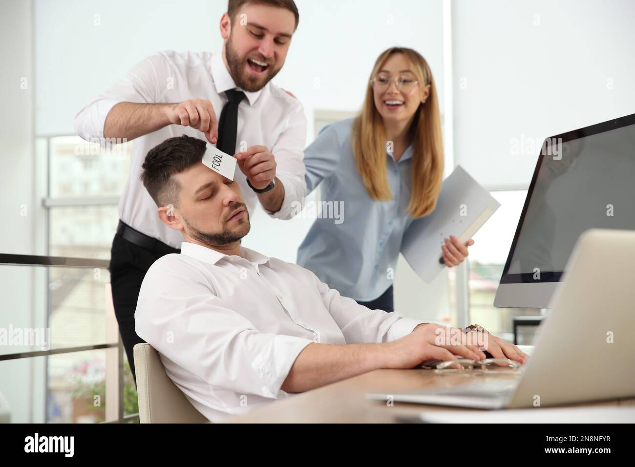 Coworkers sticking note with word Fool to colleague's face in office ...