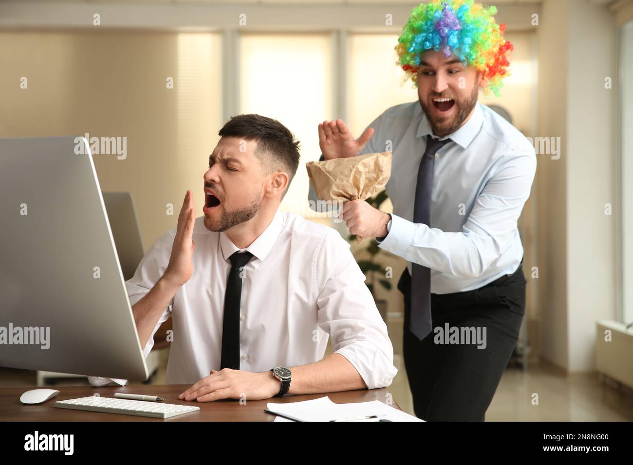 Young man popping paper bag behind his colleague in office. Funny joke ...