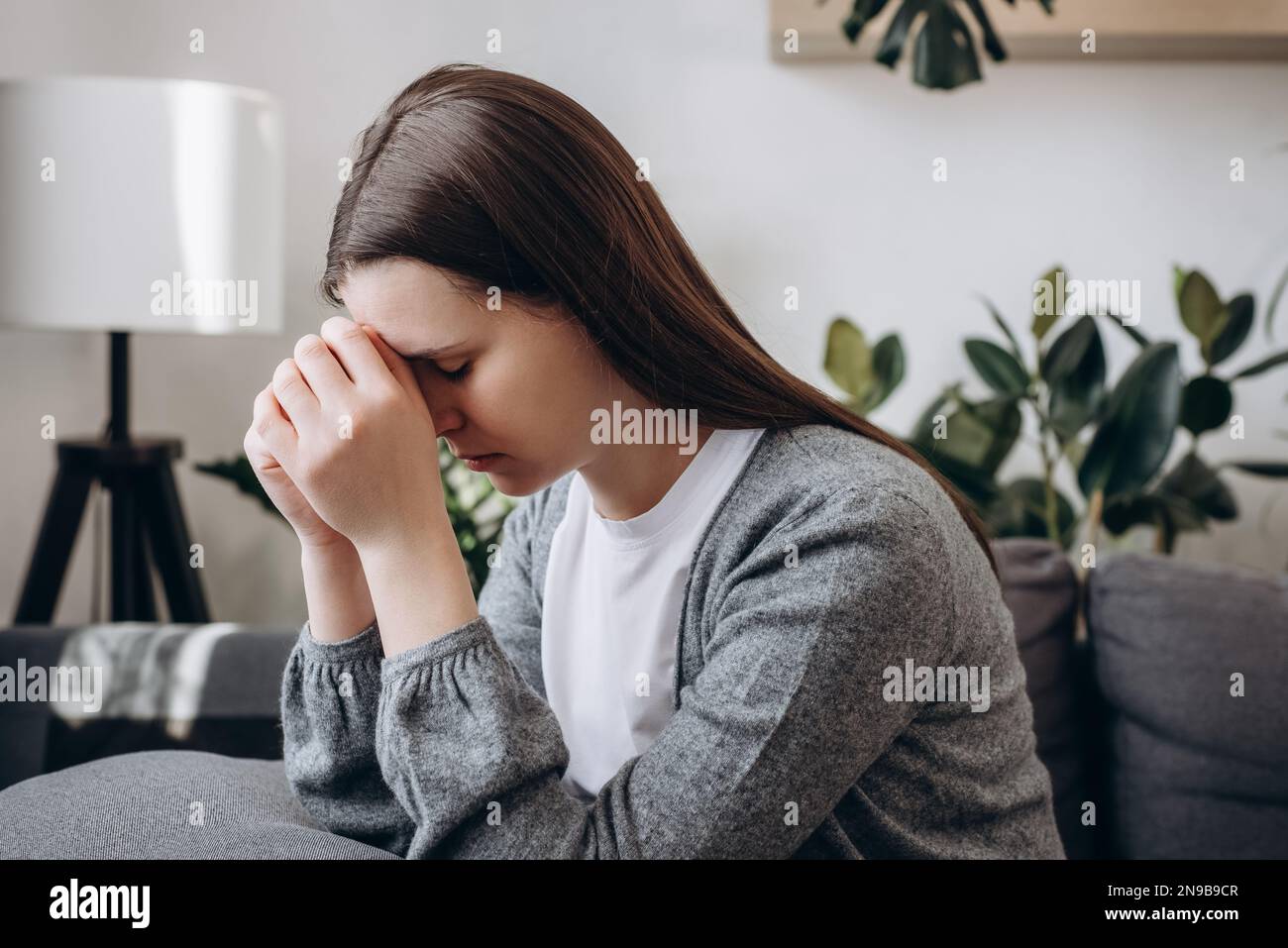 Thoughtful young female sit on couch lean forward on folded hands try ...
