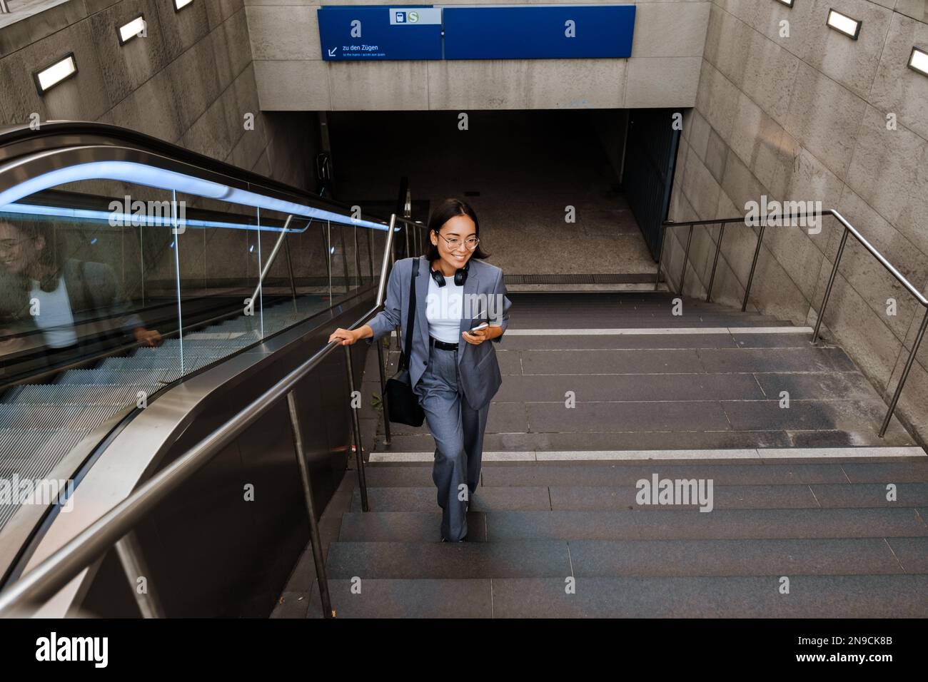 Young joyful asian woman wearing suit holding smartphone while going up ...