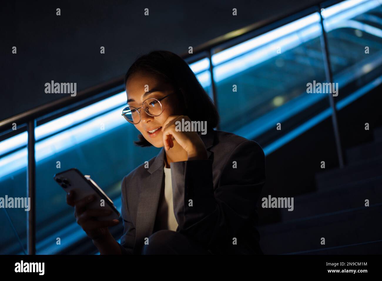 Young pensive asian woman in eyeglasses using mobile phone and drinking coffee while sitting on ...