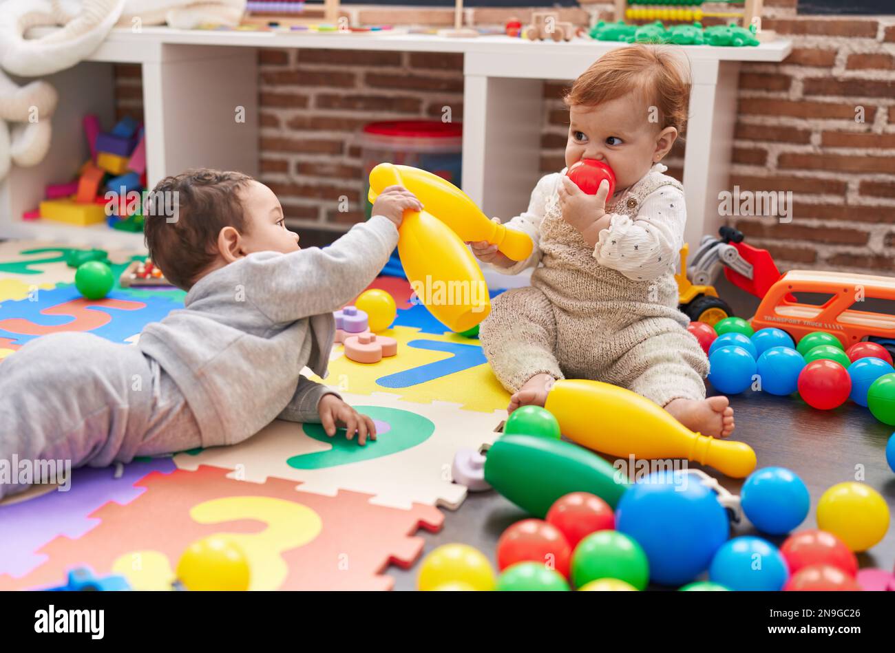 Two adorable babies playing with balls and bowling pin sitting on floor ...