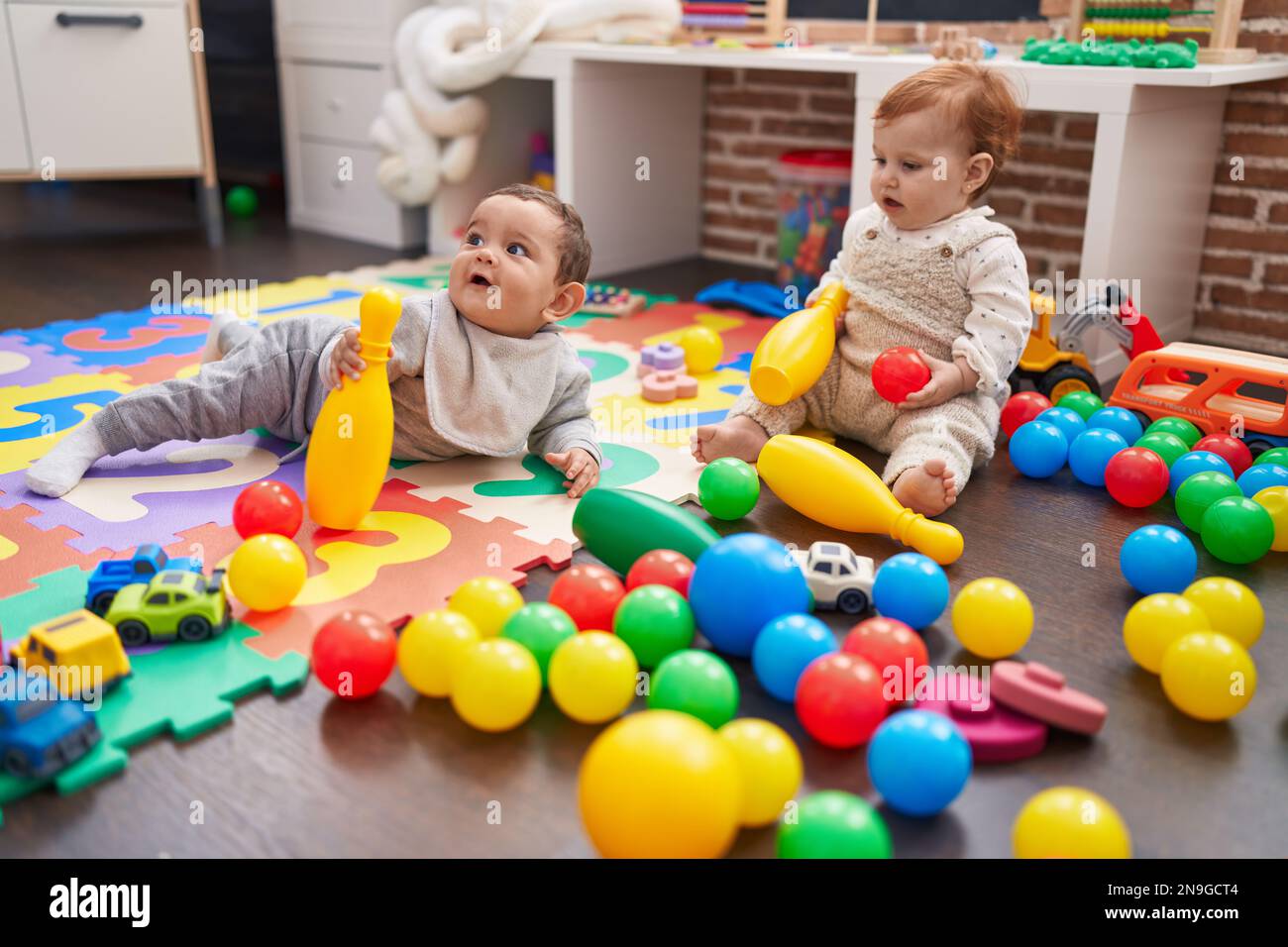 Two adorable babies playing with balls and bowling pin sitting on floor ...