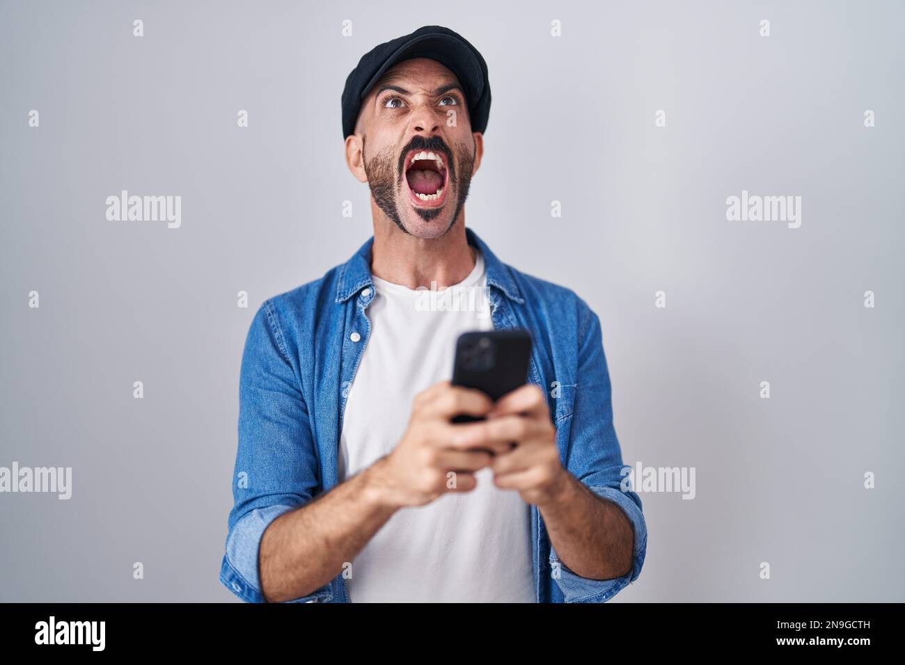 Hispanic man with beard using smartphone typing message angry and mad ...