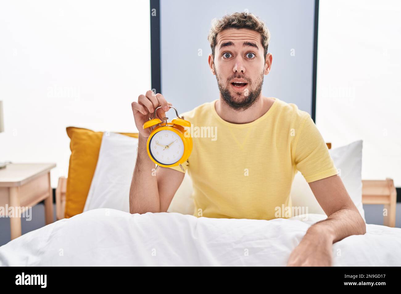 Hispanic man with beard holding alarm clock in the bed scared and ...